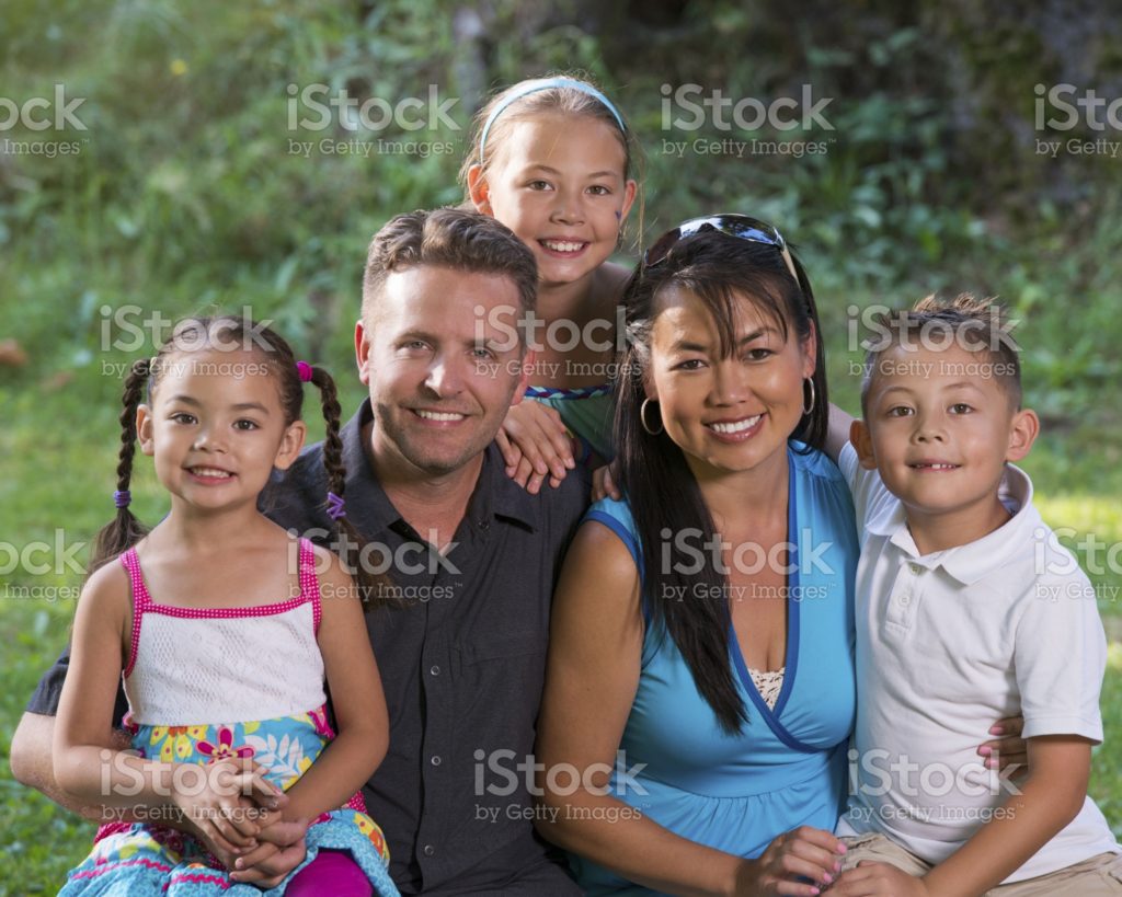 diverse family smiles / cuddling together outdoors / with grass in ...